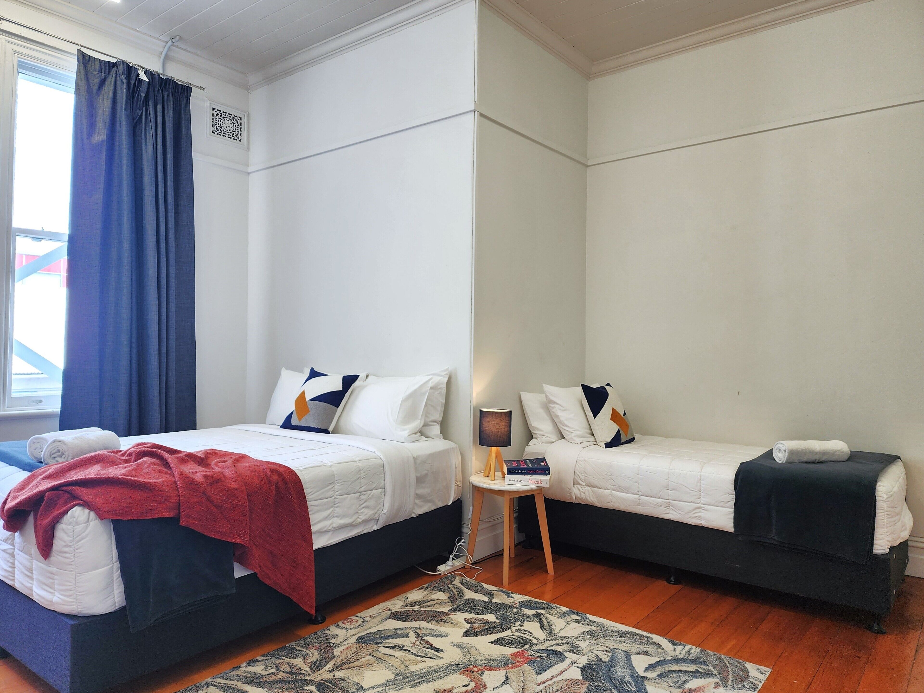 Quarto de hotel com duas camas, uma mesinha com abajur e um tapete estampado.