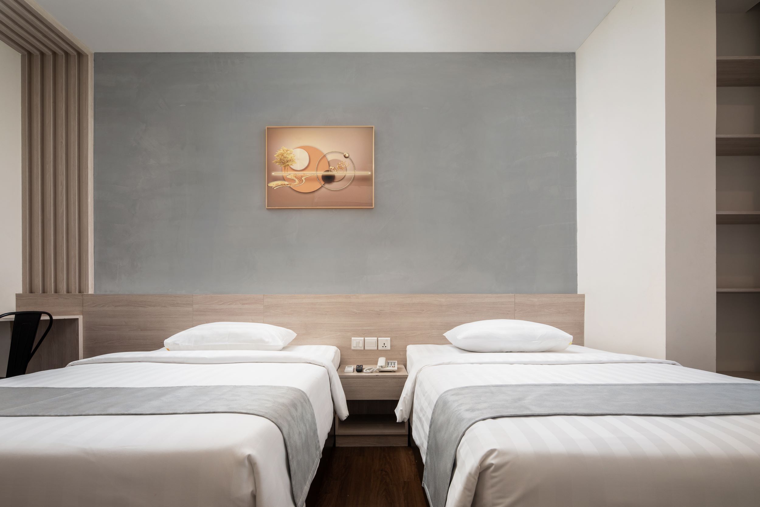 Quarto de hotel moderno com duas camas, cabeceira de madeira, mesa de cabeceira com telefone e carregador, e uma obra de arte na parede.