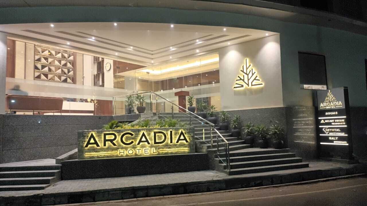 Exterior do Hotel Arcadia, com design moderno, apresentando uma entrada imponente com degraus, área de recepção e placa indicando o nome e os serviços do hotel.