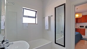 Casa de banho com banheira, chuveiro e lavatório. Uma janela permite a entrada de luz natural e há um espelho sobre o lavatório.