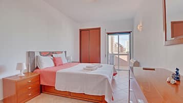 Quarto com cama, mesa de cabeceira e cômoda. Uma porta dá acesso a uma varanda com vista para prédios.