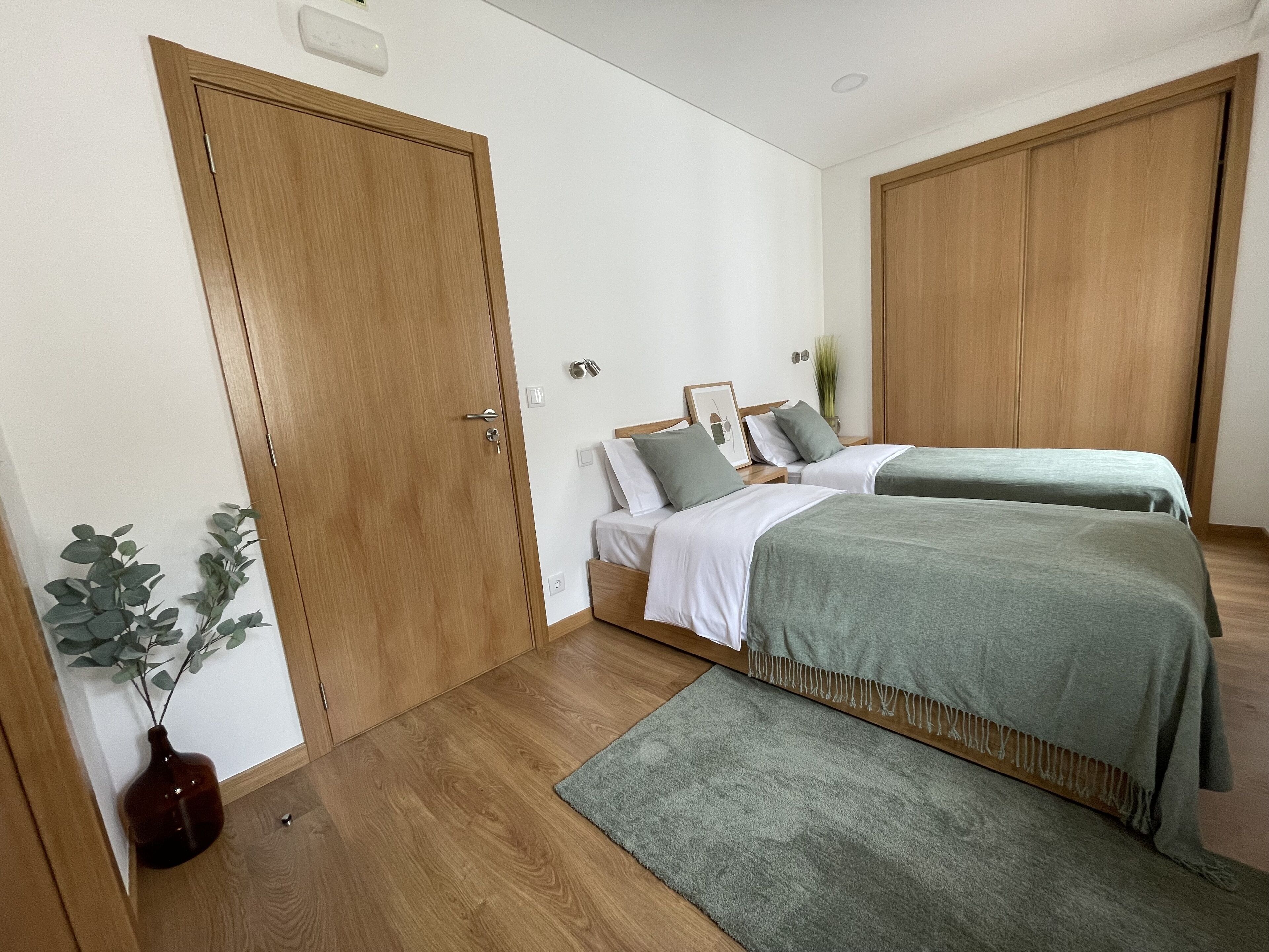 Quarto com duas camas, armários de madeira, uma planta pequena e um tapete cinza.
