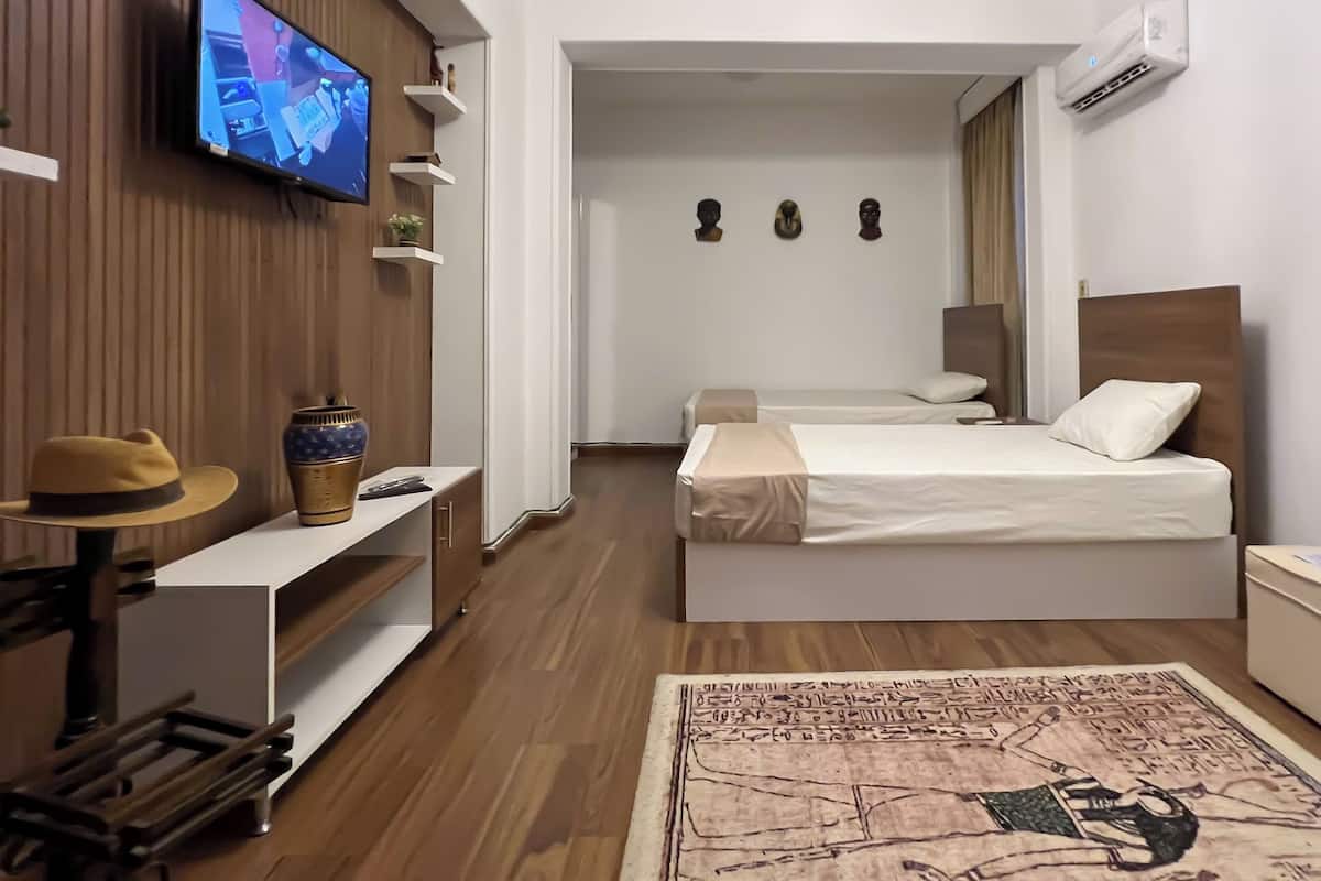 Quarto de hotel com piso de madeira, cama com colchão branco e roupa de cama bege, mesa de cabeceira pequena, TV fixada na parede e um vaso decorativo em uma prateleira.