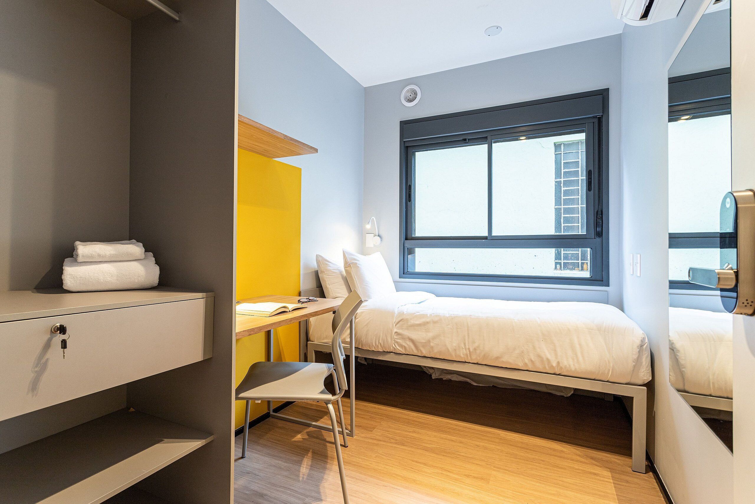 Quarto compacto com cama, escrivaninha, cadeira, guarda-roupa e janela.