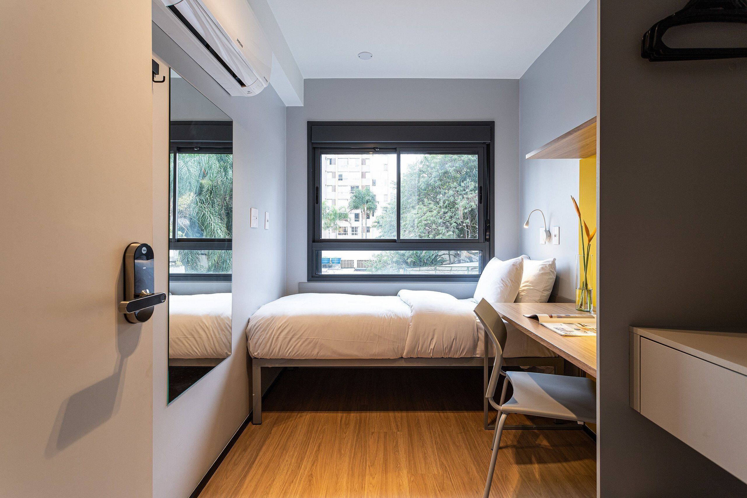 Quarto de hotel compacto com cama, escrivaninha e cadeira, uma janela com vista para prédios e árvores, e um aparelho de ar condicionado.