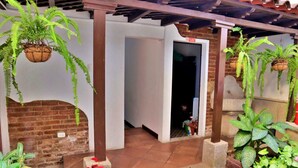 Área externa coberta com samambaias penduradas, parede de tijolos e piso ladrilhado.