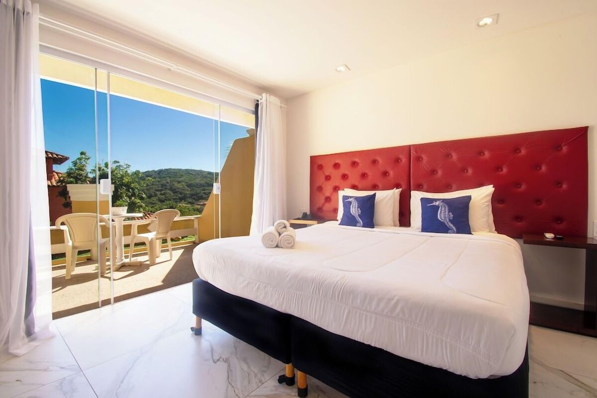 Quarto de hotel com uma cama grande, cabeceira vermelha e varanda com mesa e cadeiras.