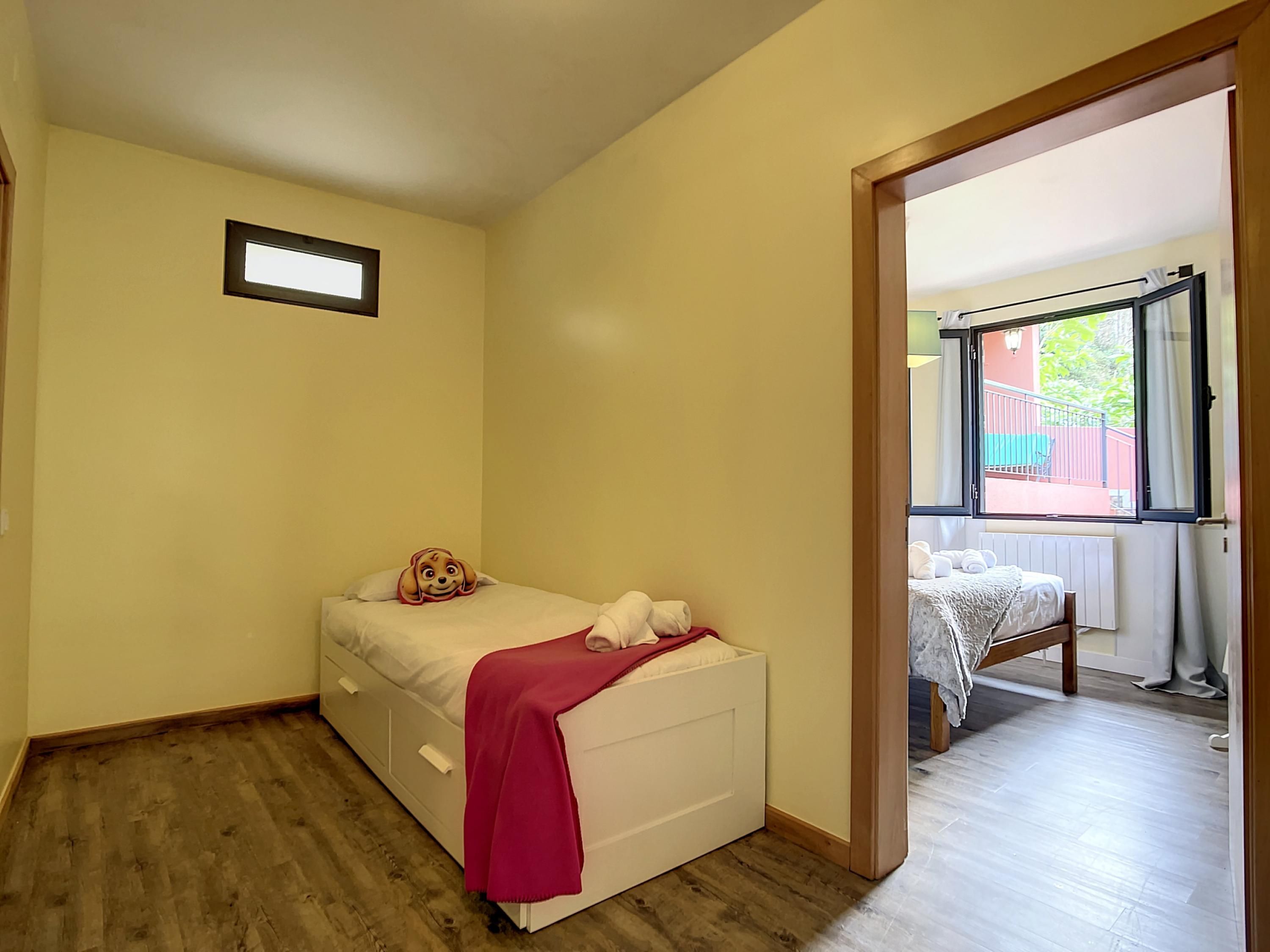 Quarto pequeno com uma cama de solteiro, piso de madeira, um cobertor vermelho e janela com vista para uma varanda.