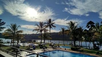 Piscina com espreguiçadeiras e guarda-sóis, cercada por palmeiras e vista para a água.