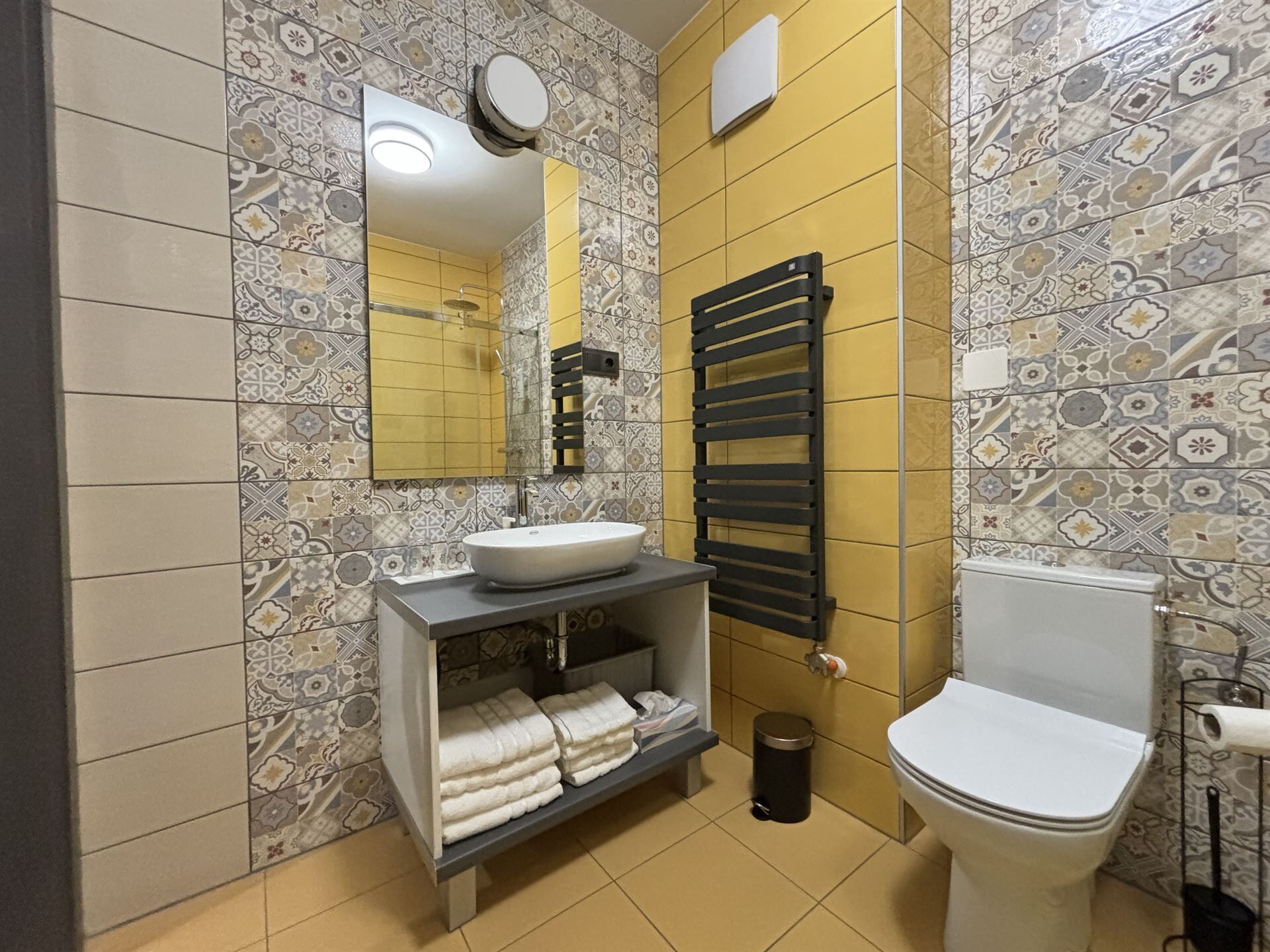 Casa de banho com lavatório, espelho, porta-toalhas, sanita e azulejos estampados.