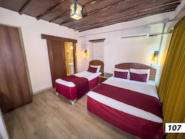 Quarto de hotel com duas camas, piso de madeira, teto de madeira, um espelho, uma pia e uma janela.