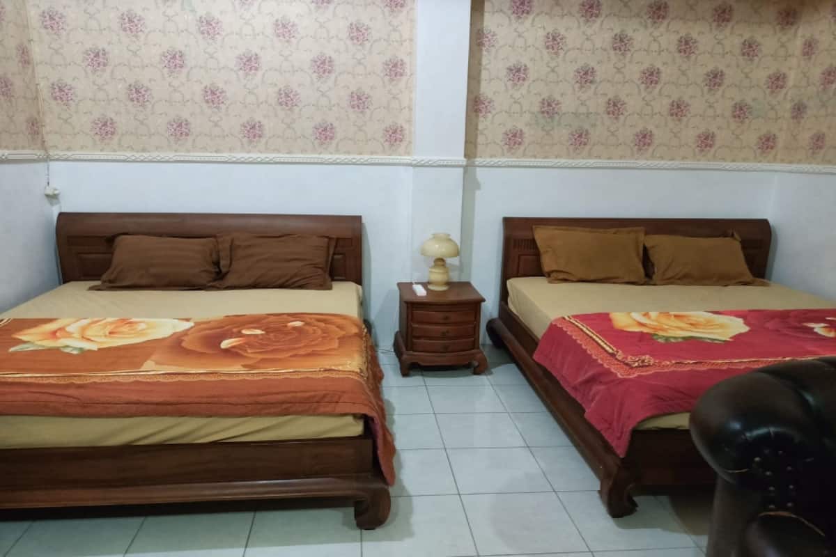Quarto com duas camas de madeira, cada uma com colcha florida e travesseiros marrons. Pequeno criado-mudo de madeira entre as camas, com uma luminária sobre ele. As paredes têm um padrão de papel de parede floral.