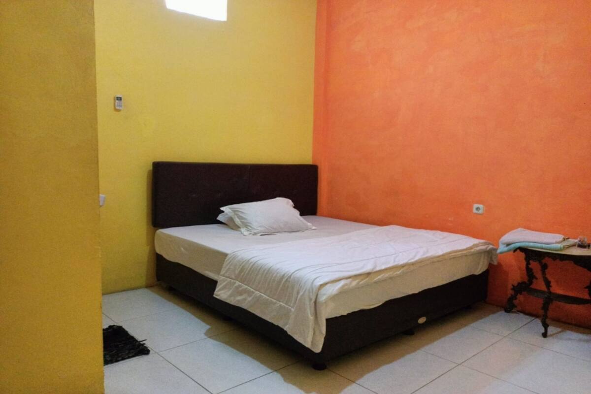 Quarto pequeno com uma cama de solteiro, cabeceira escura, roupa de cama branca e uma pequena mesa com uma toalha.