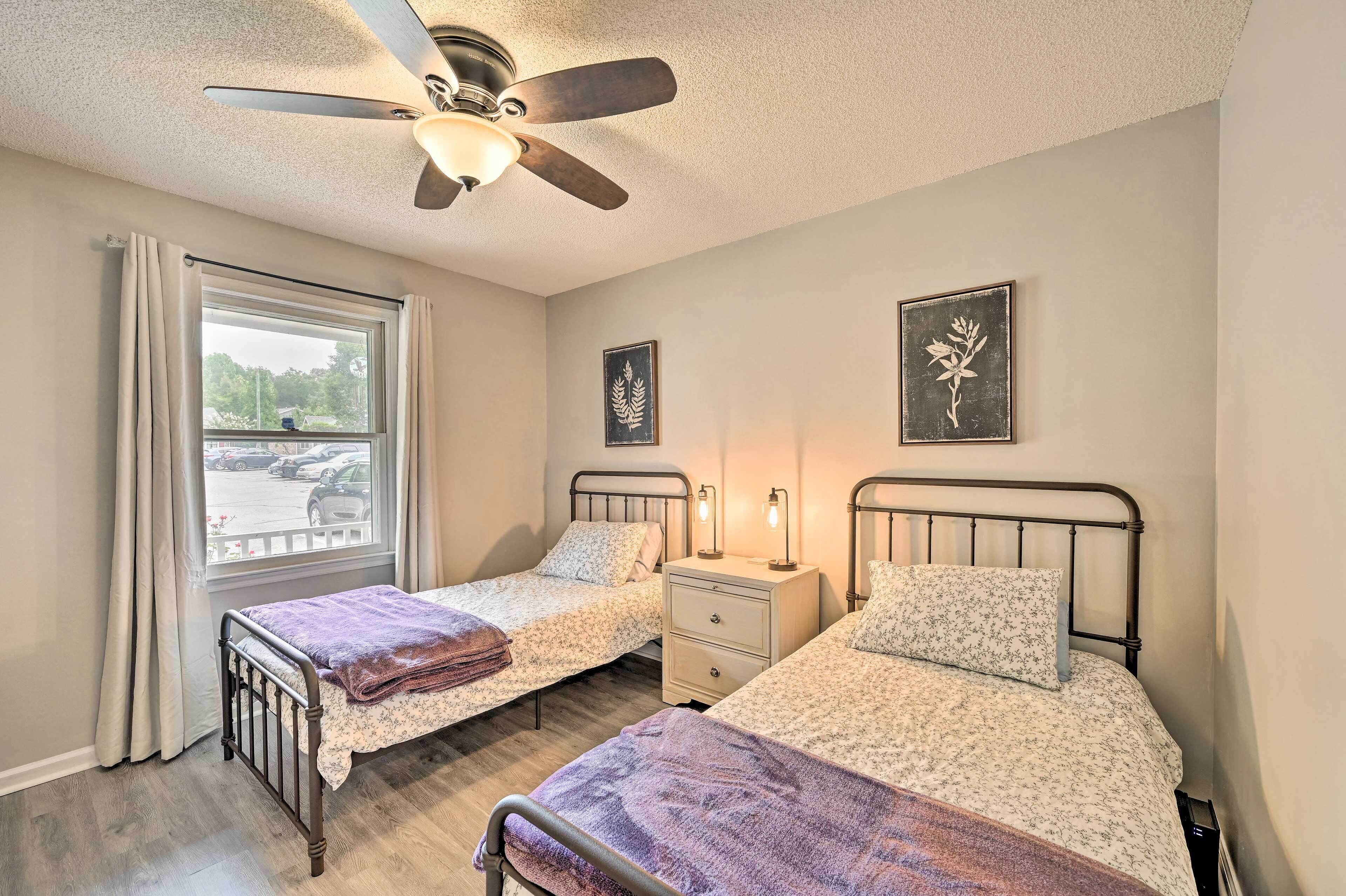 Quarto com duas camas de solteiro, um ventilador de teto, uma janela com cortinas e duas luminárias fixadas na parede.