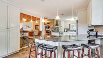 Uma cozinha ampla com armários brancos, uma ilha grande com banquetas, iluminação pendente e vista para a área de refeições.