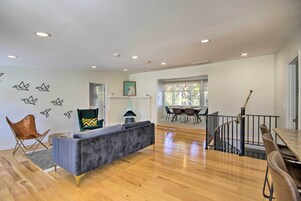 Uma sala de estar ampla, com sofá cinzento, piso de madeira, mesa de jantar com cadeiras, escada e vista para o exterior.