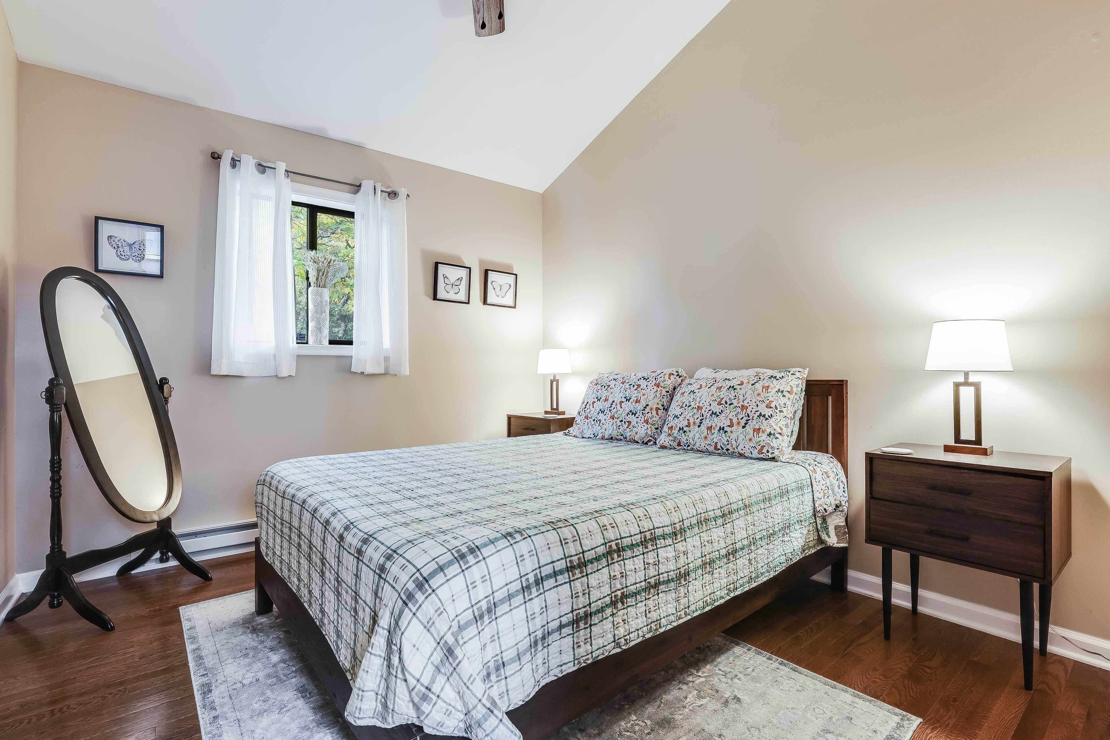 Quarto com uma cama, duas mesas de cabeceira com luminárias, um espelho de corpo inteiro e quadros nas paredes.