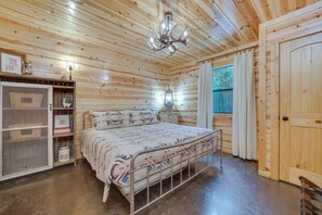 Quarto de cabana de madeira com cama de metal, guarda-roupa de madeira, um candeeiro e janela com cortinas.