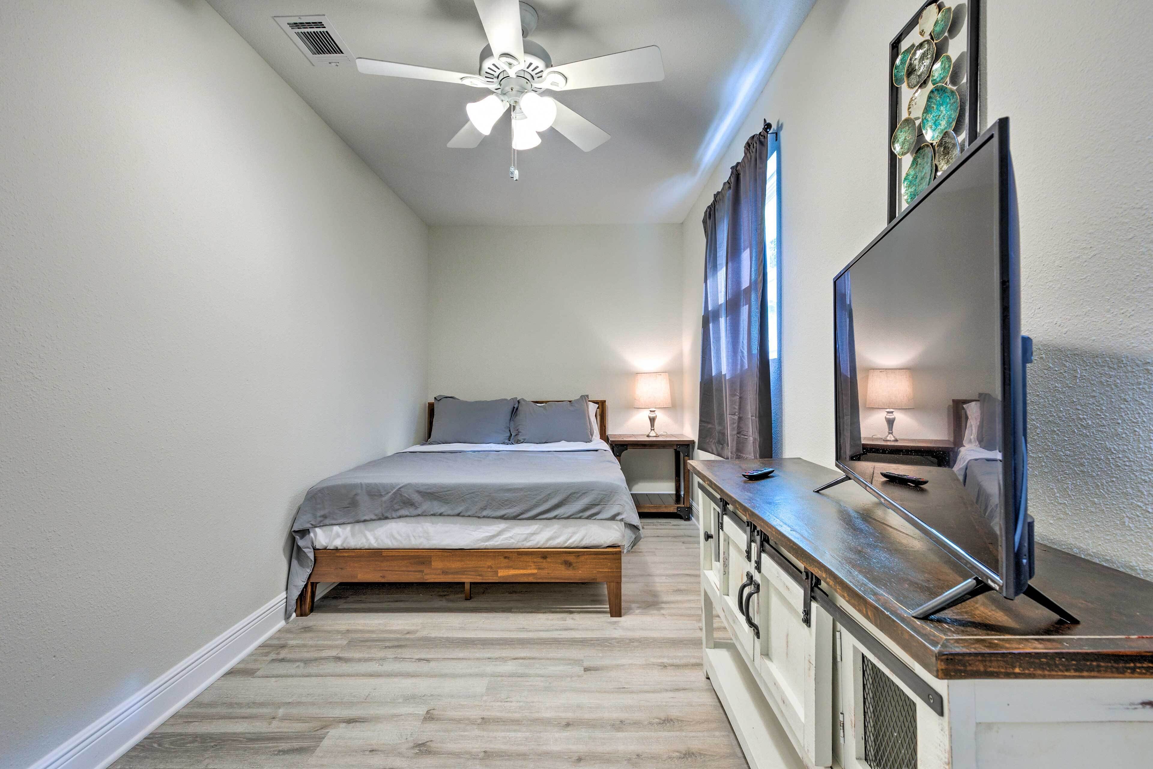 Quarto com cama, ventilador de teto, televisão e mesa de cabeceira com luminária.