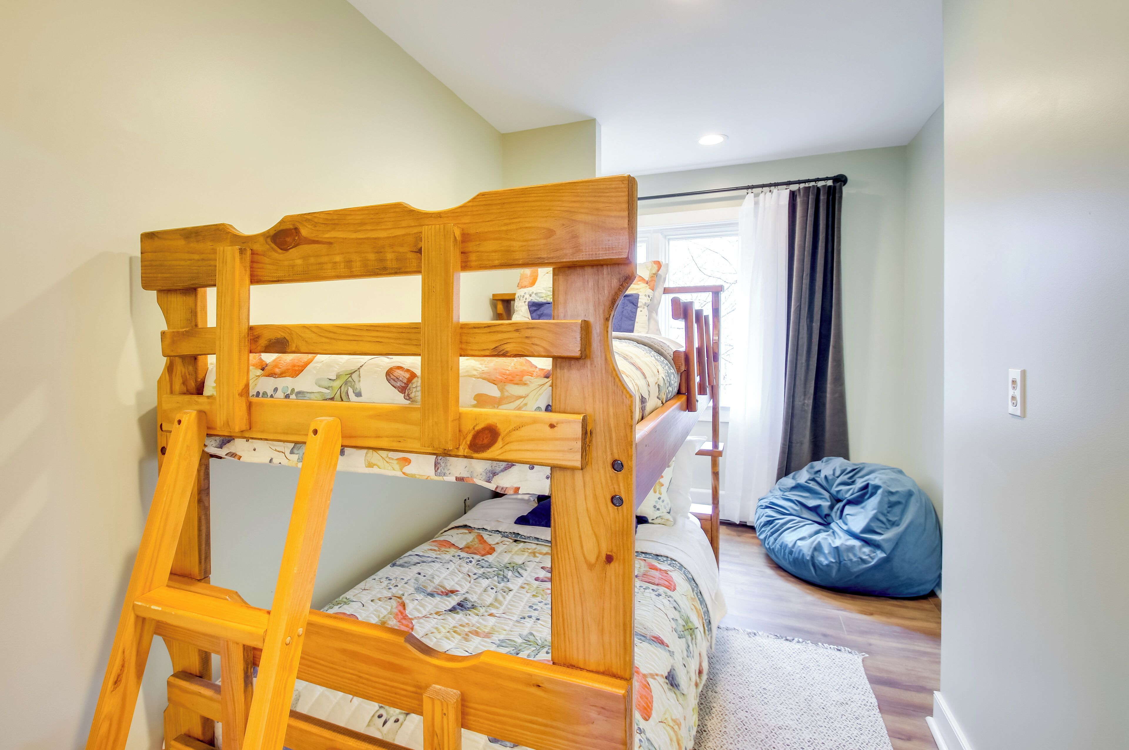Uma cama de beliche com estrutura de madeira e roupa de cama floral, uma poltrona de couro azul e uma janela com cortinas.