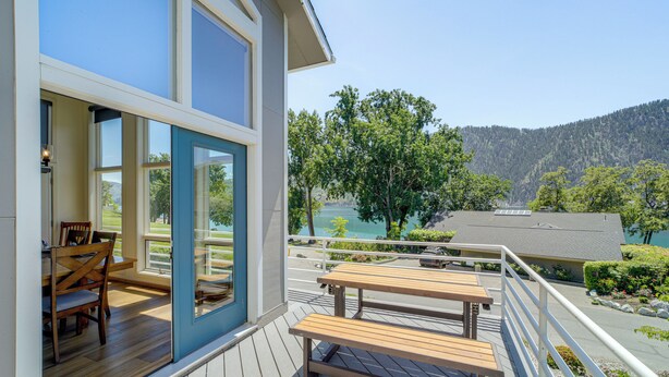 Uma casa moderna com um deck, uma área de refeições com cadeiras e uma mesa, e vista para um lago e uma floresta.