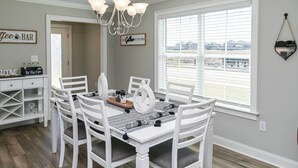 Uma sala de jantar com uma mesa branca posta para quatro pessoas, cadeiras brancas, um lustre e vista através de janelas amplas.