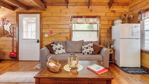 Interior acolhedor de uma cabana com piso de madeira, um sofá com almofadas estampadas, uma mesa de centro com um cesto e um candeeiro, e uma geladeira com objetos decorativos.