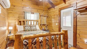 Quarto de cabana de madeira com cama, mesa de cabeceira, janela com cortinas e porta.