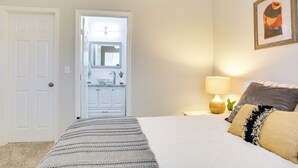 Quarto com cama, luminária de cabeceira, quadro emoldurado e porta para banheiro com espelho e pia.