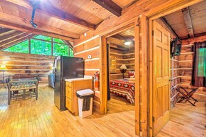 Interior de cabana de tronco com cozinha, mesa de jantar e um quarto aconchegante com colcha estampada.