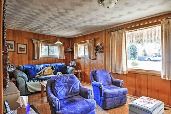 Um quarto acolhedor com paredes de madeira, um sofá azul, duas poltronas, uma mesa pequena e uma janela com cortinas.