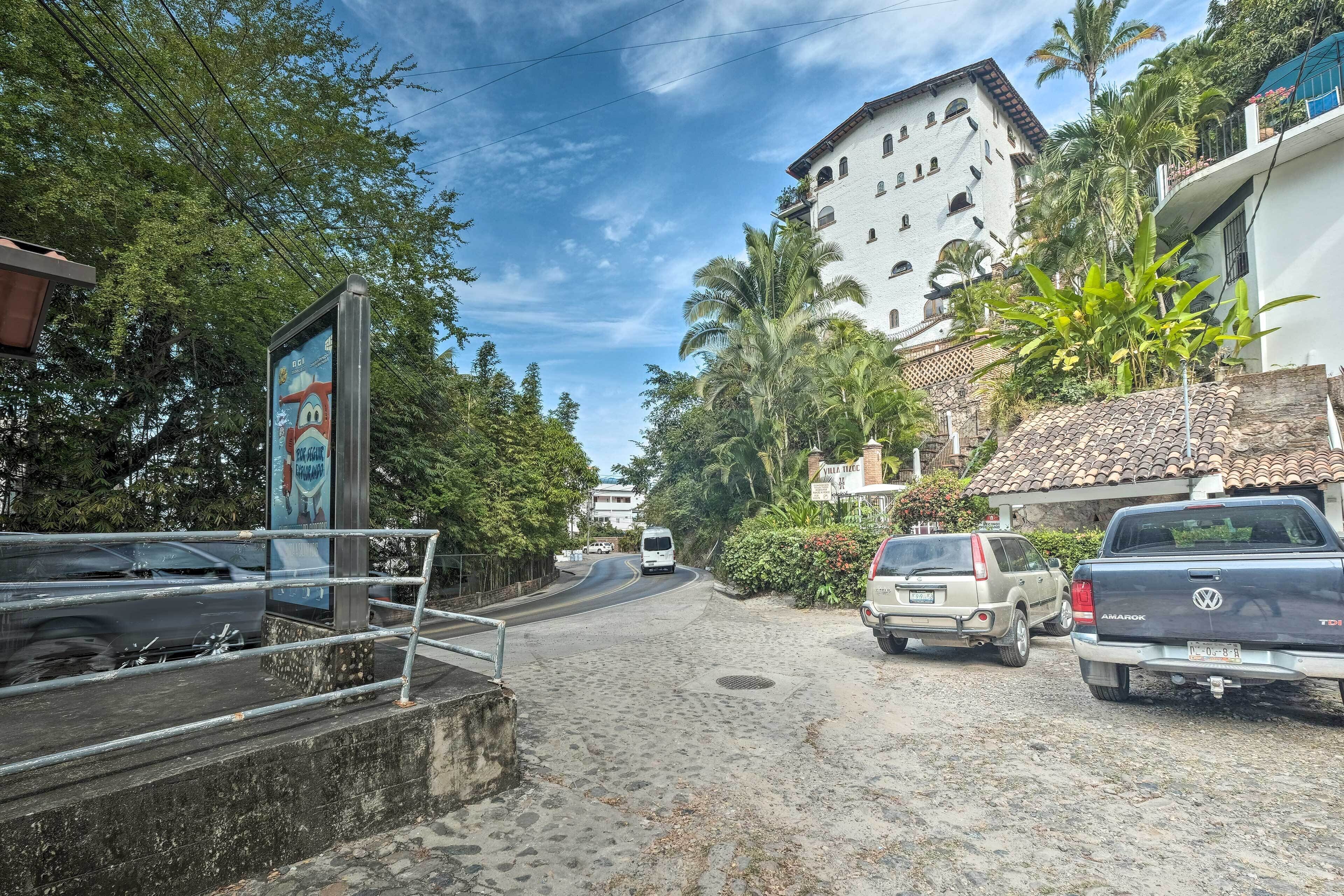 Uma rua de paralelepípedos com carros estacionados, um edifício branco com várias janelas e vegetação exuberante.