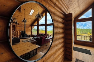 Interior de uma cabana de madeira com um espelho grande refletindo o ambiente, lareira, uma área de estar aconchegante e vista para a floresta através de amplas janelas.