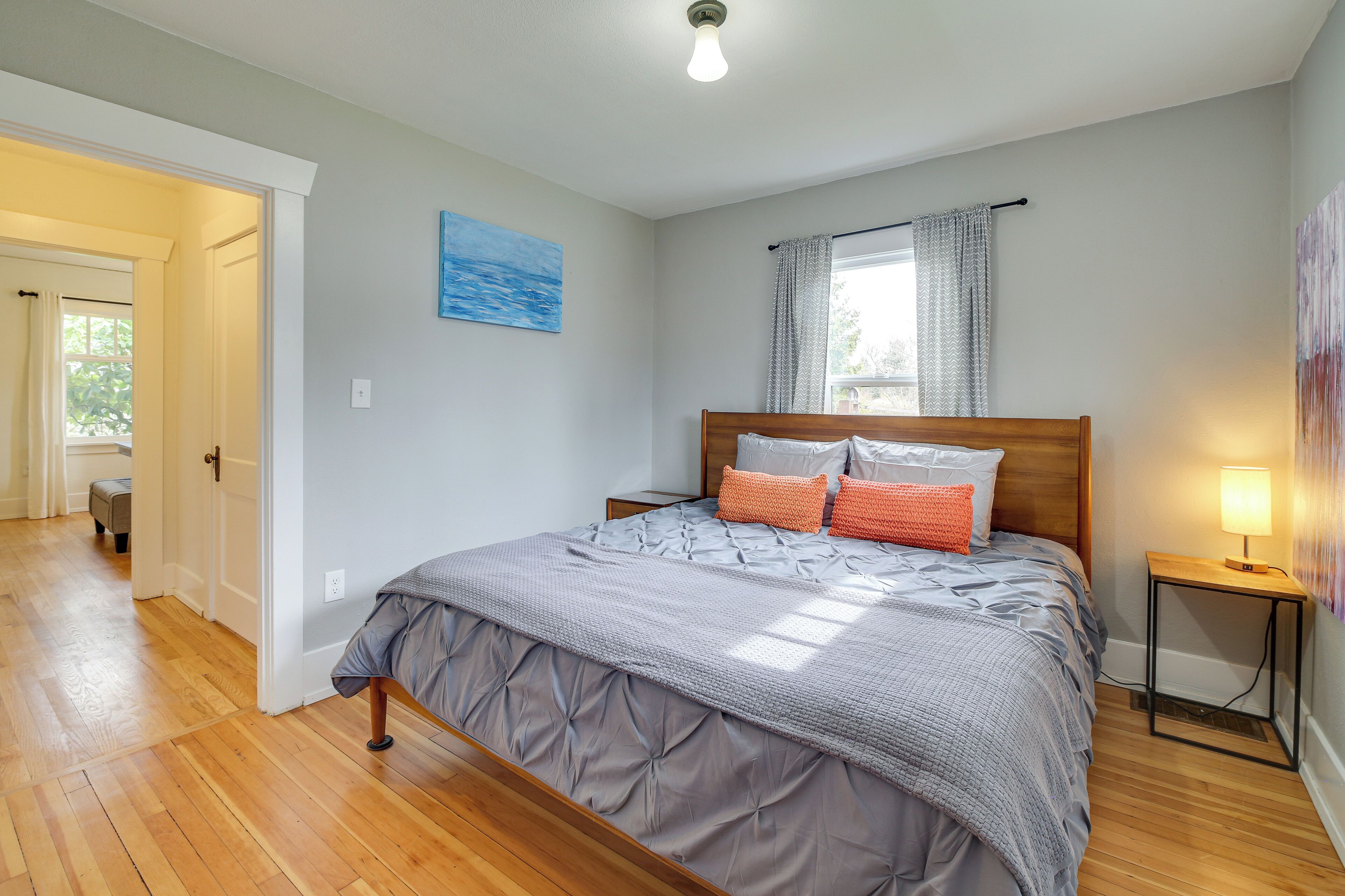 Quarto com cama de madeira, mesa de cabeceira, abajur, janela com cortinas e um quadro na parede.