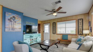 Uma sala de estar com televisão, ventilador de teto e um letreiro que diz 'BEACH HOUSE'.