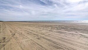 Uma extensa praia de areia com marcas de pneus e uma vista distante do oceano sob um céu nublado.