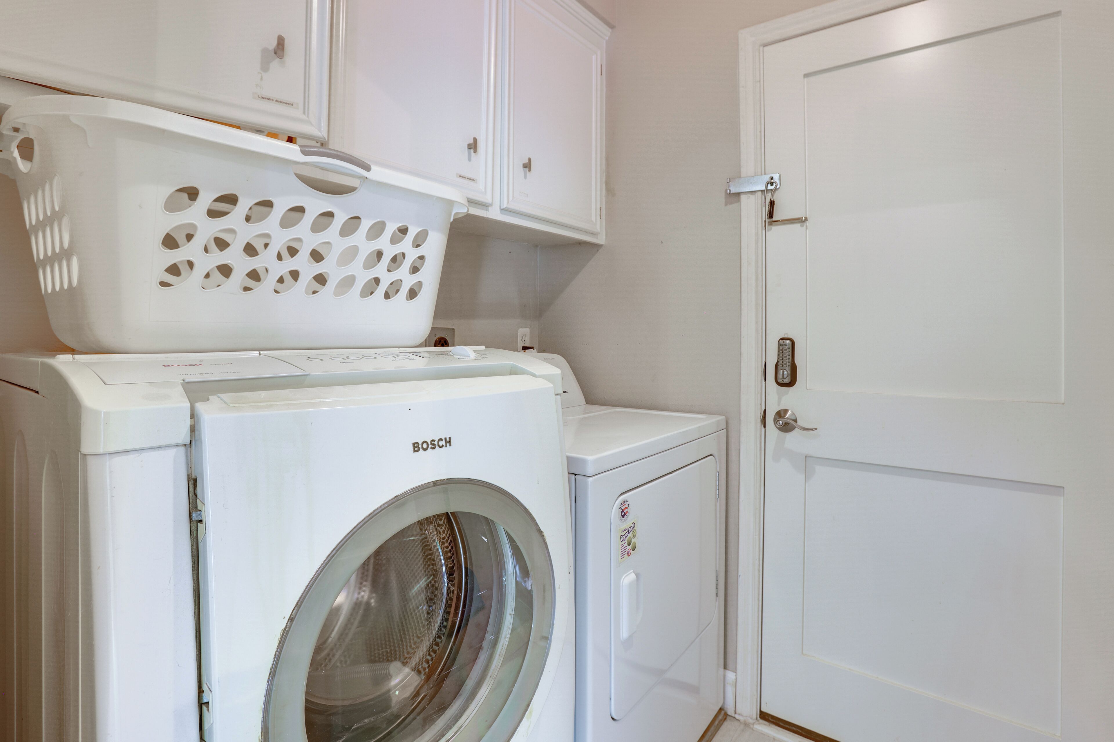 Uma lavanderia com máquina de lavar frontal, secadora, cesto de roupa branco e porta com puxador.