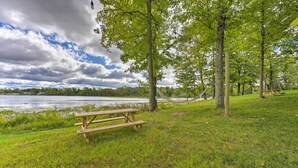 Uma mesa de piquenique e uma rede em área gramada, com árvores e um lago ao fundo.