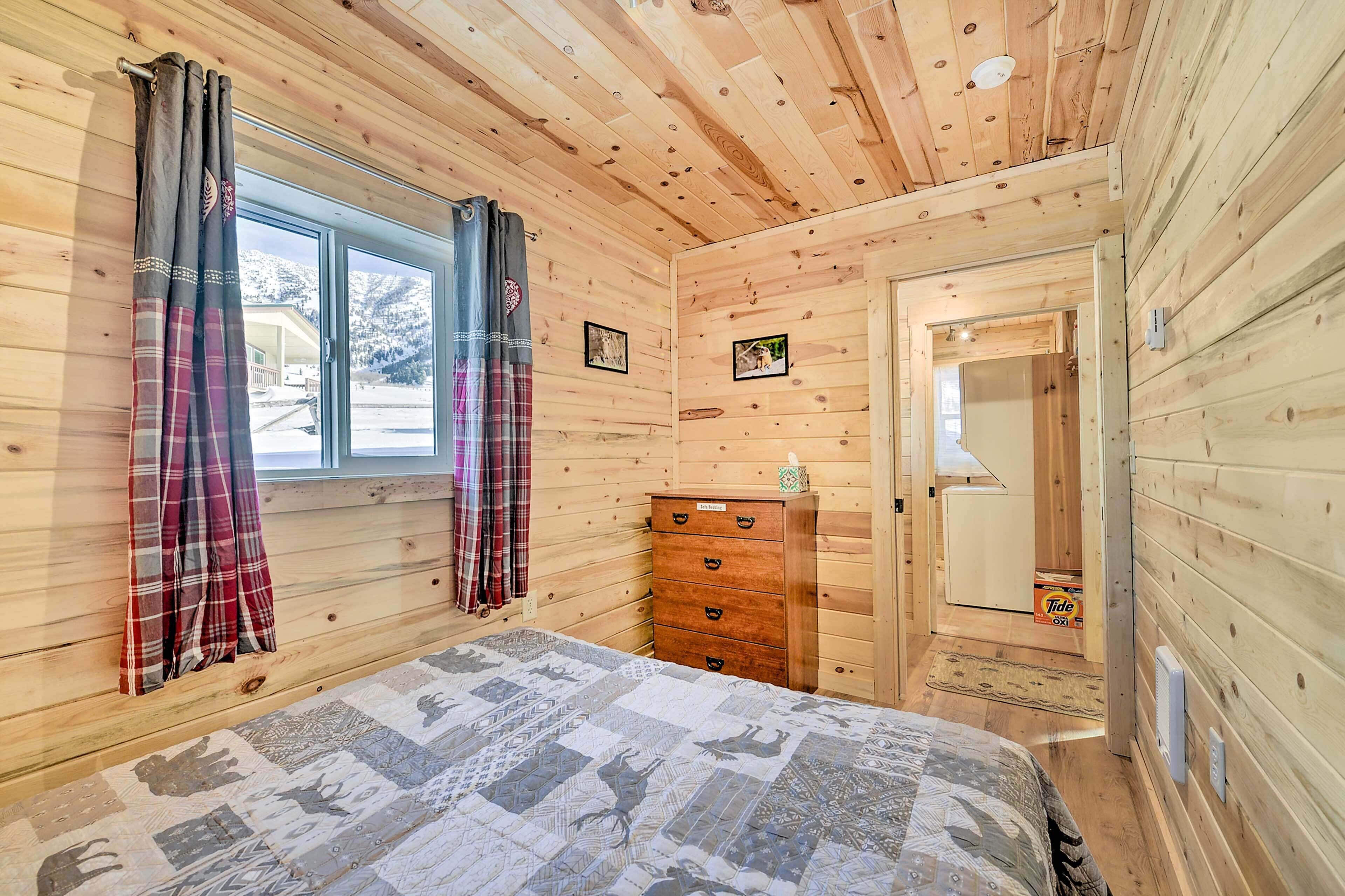 Quarto de cabana de madeira com cama, penteadeira, janela com cortinas e porta que dá para outro cômodo.