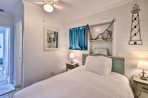 Quarto com cama, mesa de cabeceira, abajur, ventilador de teto, janela com cortinas e prateleira na parede com objetos decorativos.