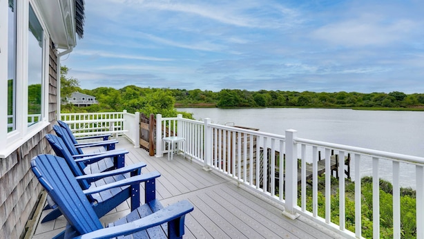 Um deck de madeira com cadeiras Adirondack azuis, um corrimão branco e vista para um lago.