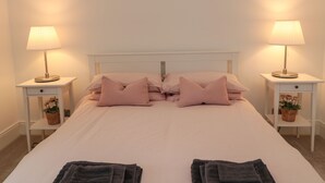 Cama bem arrumada com duas mesas de cabeceira, cada uma com uma luminária e uma plantinha.