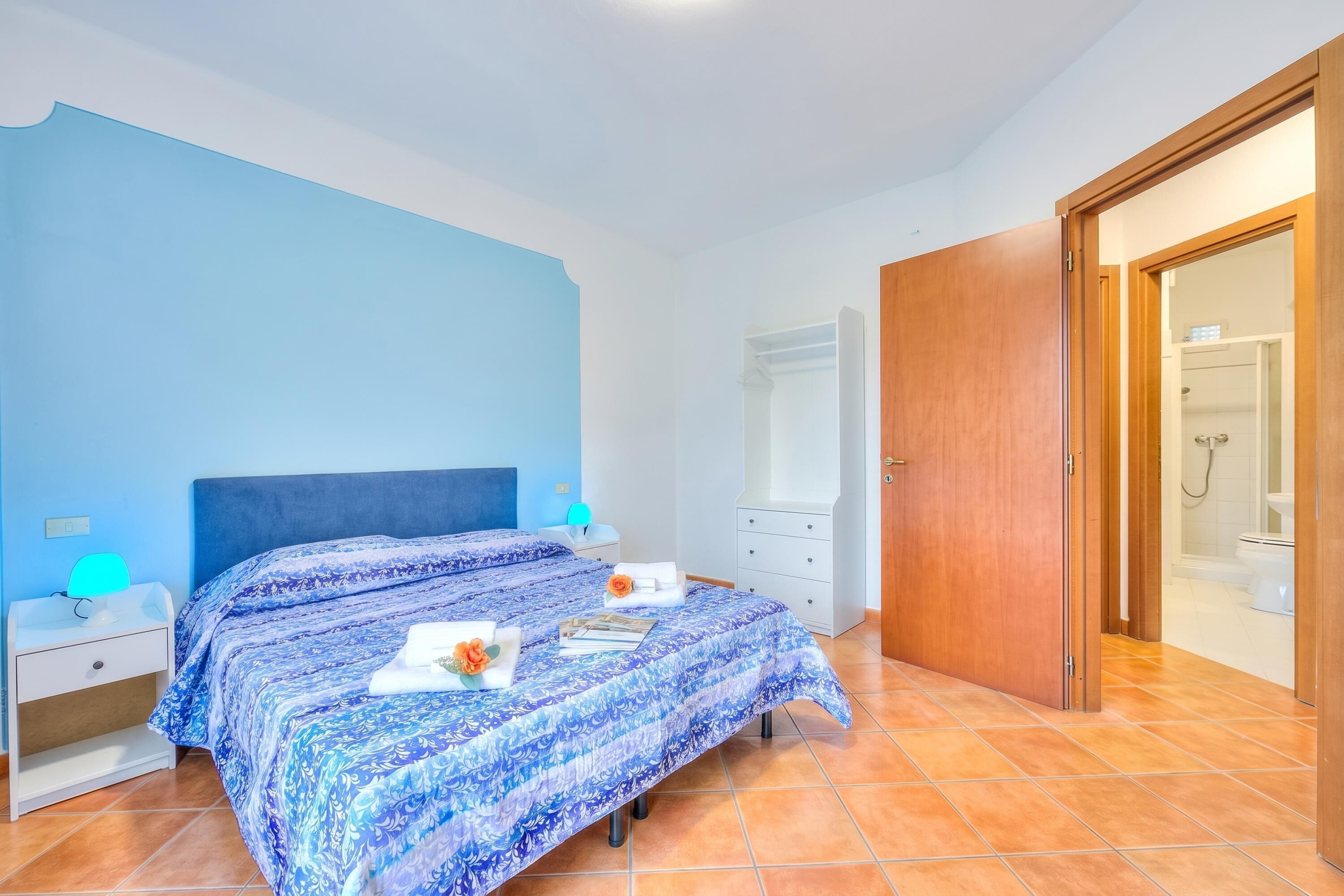 Quarto com cama, mesas de cabeceira, guarda-roupa e um banheiro visível por uma porta aberta.