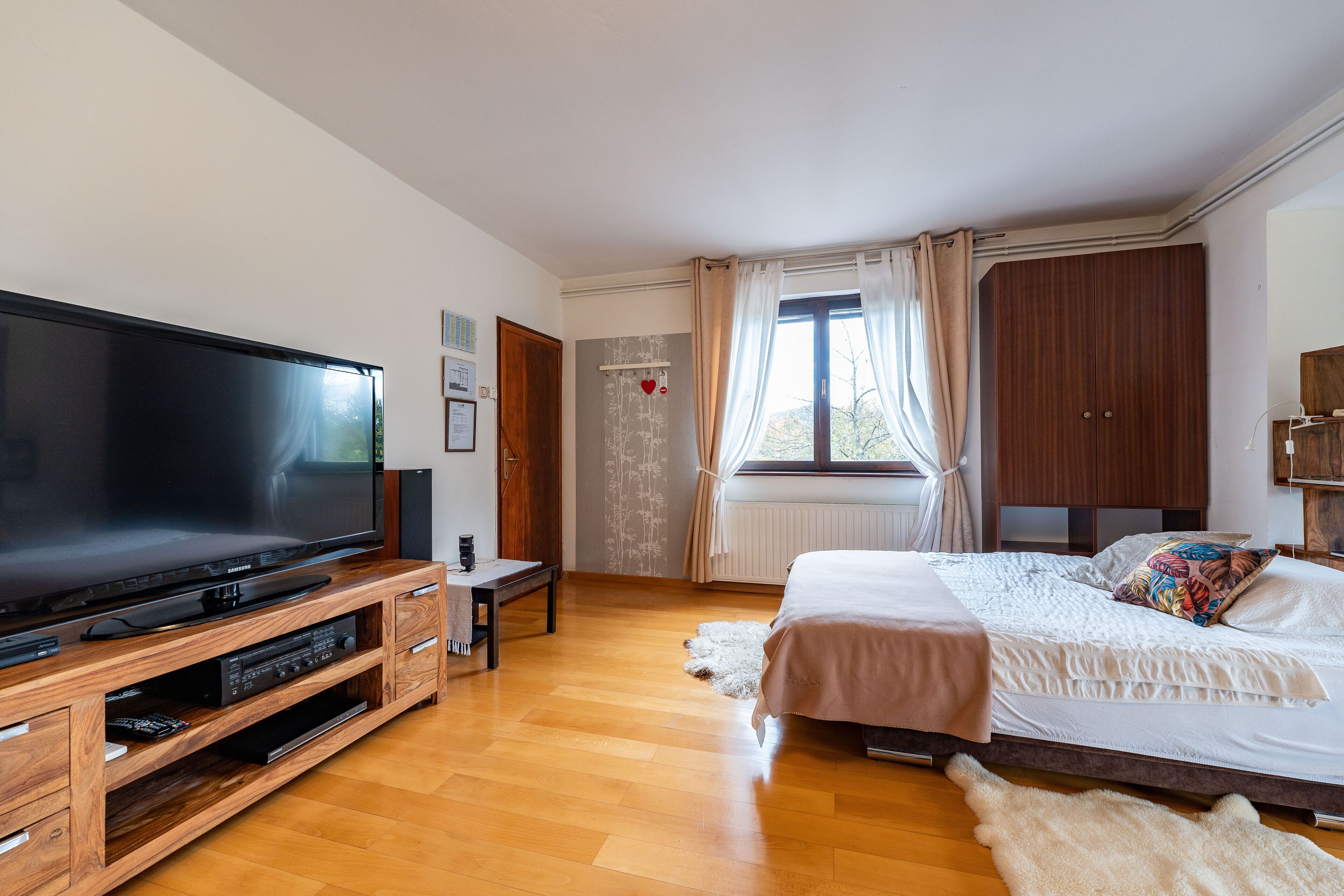 Quarto com televisão de tela grande, móveis de madeira, cama com edredom branco e janela com cortinas.