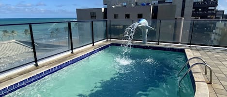 Uma piscina no terraço com água azul cristalina, rodeada por um guarda-corpo de vidro e um deck revestido de azulejos.