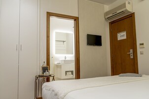 Quarto de hotel com uma cama, uma pequena mesa com uma garrafa e uma placa, uma TV presa na parede e uma porta com uma placa.