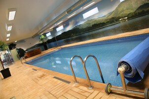 Piscina interior com deck de madeira, guarda-corpos de metal e um mural com paisagem de lago e montanhas na parede.