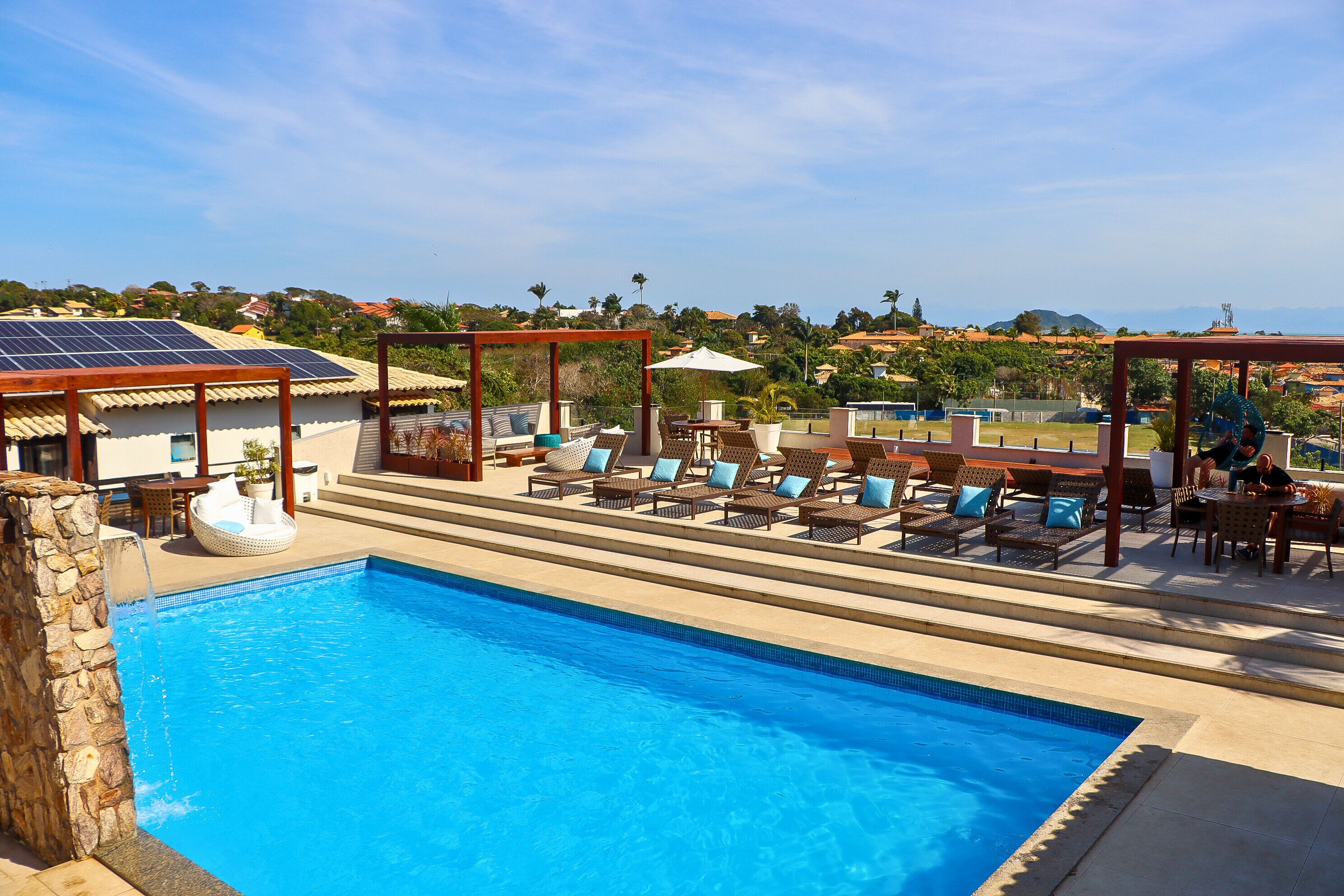 Piscina com espreguiçadeiras e guarda-sóis, cercada por um muro de pedra e um pergolado.