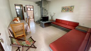Uma sala de estar com um sofá vermelho, uma mesa de jantar com cadeiras, um rack para televisão e uma escada.