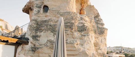 Um edifício de pedra com fachada singular que se assemelha a rocha, área de estar externa com cadeiras de vime e uma mesa, e vista para a paisagem circundante.
